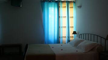 Quarto de hotel com cama, luminária de cabeceira e janela com cortinas azuis.