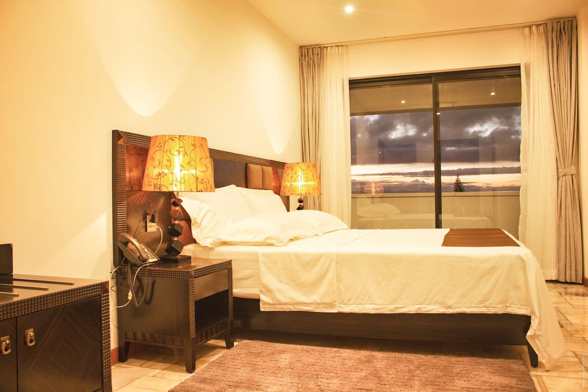 Quarto de hotel com cama, abajures, mesa de cabeceira, telefone, janela com cortinas e vista do céu.