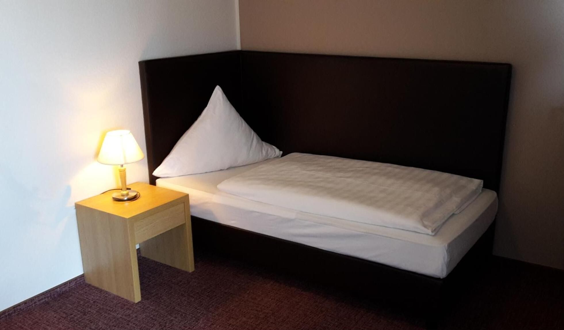 Quarto de hotel com uma cama de solteiro, mesa de cabeceira com abajur e cabeceira escura.