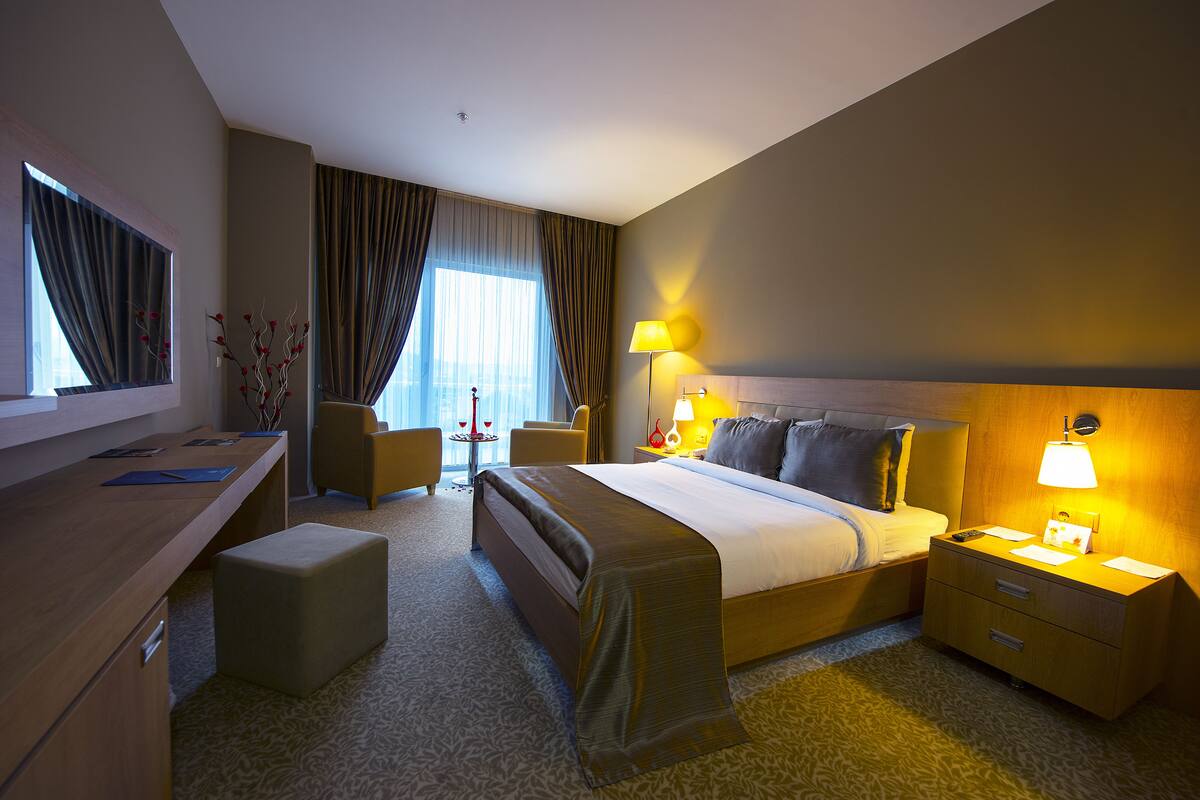 Quarto de hotel com cama, mesa de cabeceira, abajur, escrivaninha, cadeira e uma pequena mesa com um vaso.