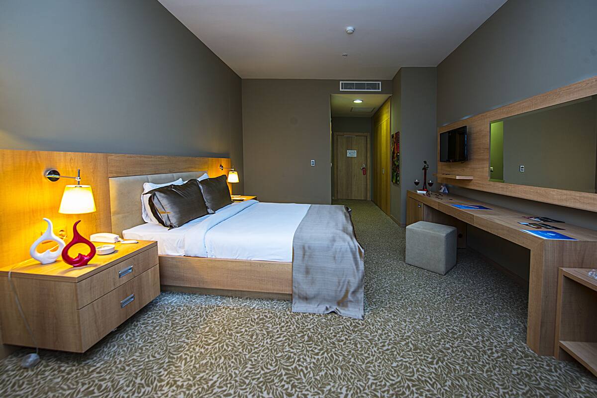 Quarto de hotel com cama, mesas de cabeceira, escrivaninha, cadeira, espelho e piso coberto por carpete.