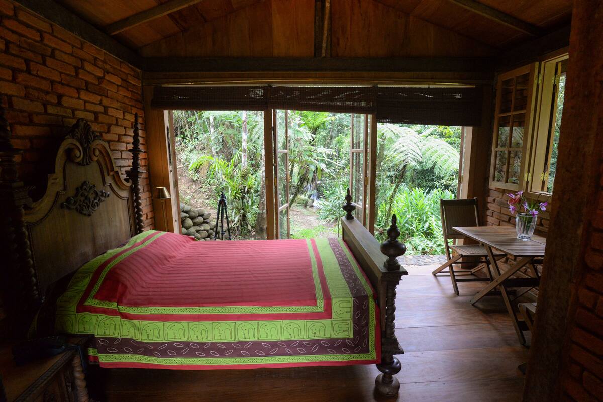 Cama de madeira com colcha vermelha e verde em quarto com parede de tijolos e janelas amplas.
