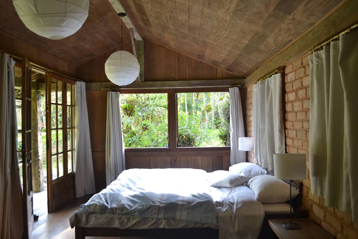 Quarto com teto de madeira, janelas amplas, cama com roupa de cama branca e luminária de cabeceira.
