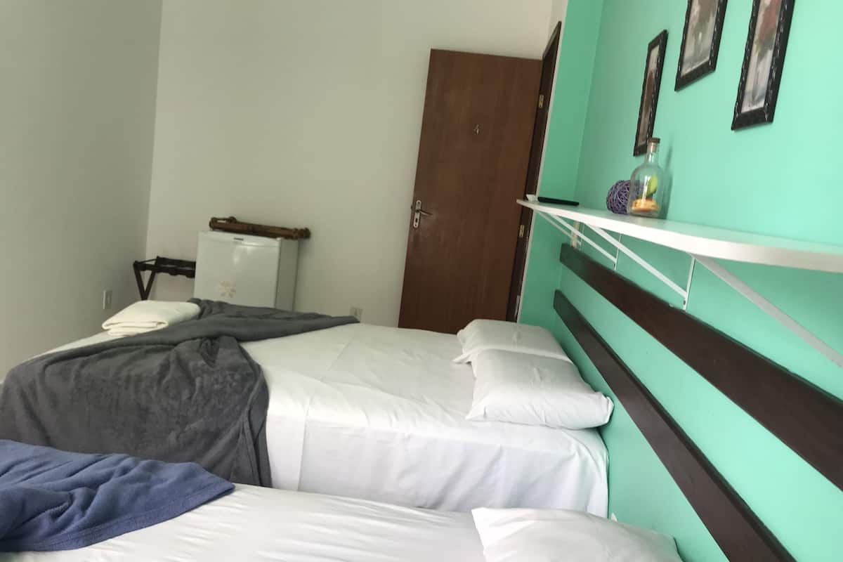 Quarto com duas camas, um guarda-roupa de madeira, uma mesinha com uma garrafa e uma prateleira na parede.