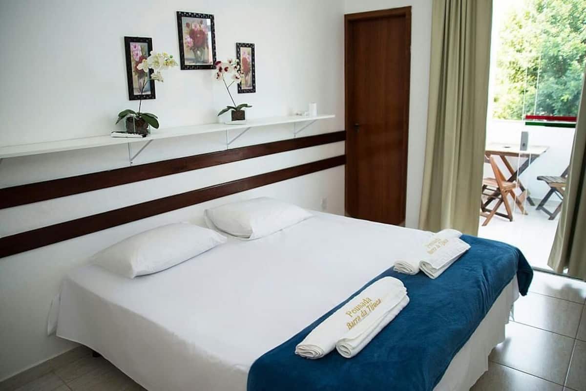 Quarto de hotel com uma cama, paredes brancas, cabeceira de madeira, uma pequena prateleira com quadros e uma porta para uma varanda.