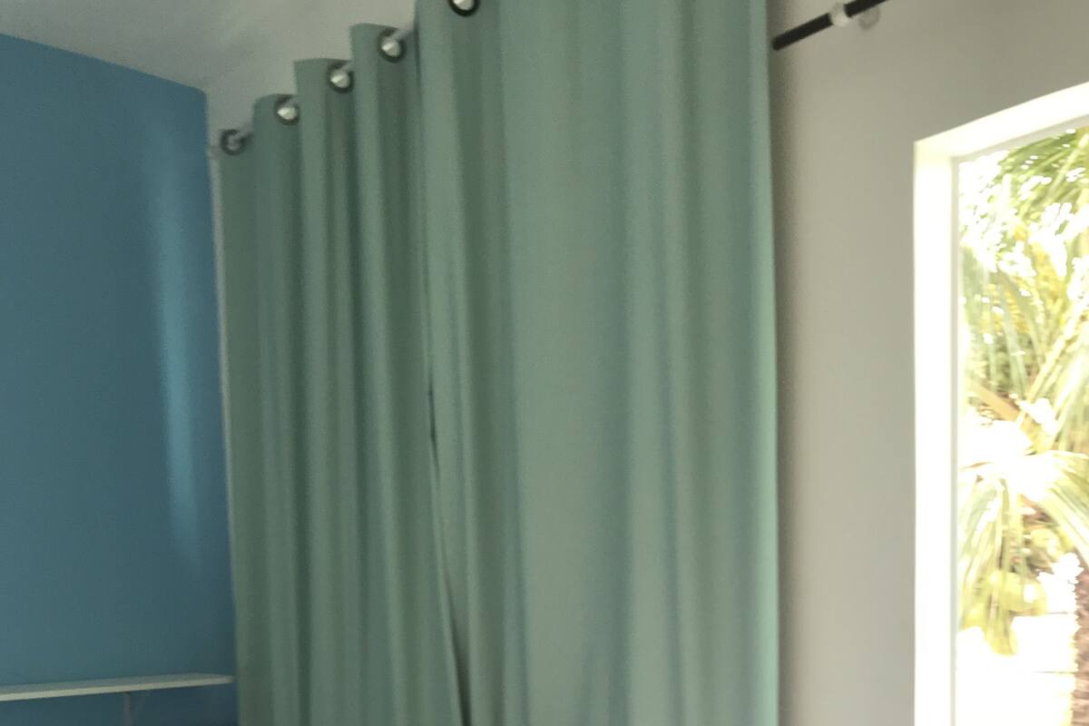 Uma cama de solteiro com colchão branco e manta vermelha, parede azul e janela com cortinas.