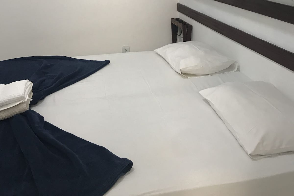 Cama bem arrumada com roupa de cama branca e azul marinho, uma prateleira na parede acima e um quadro de uma pintura de praia na parede ao lado.