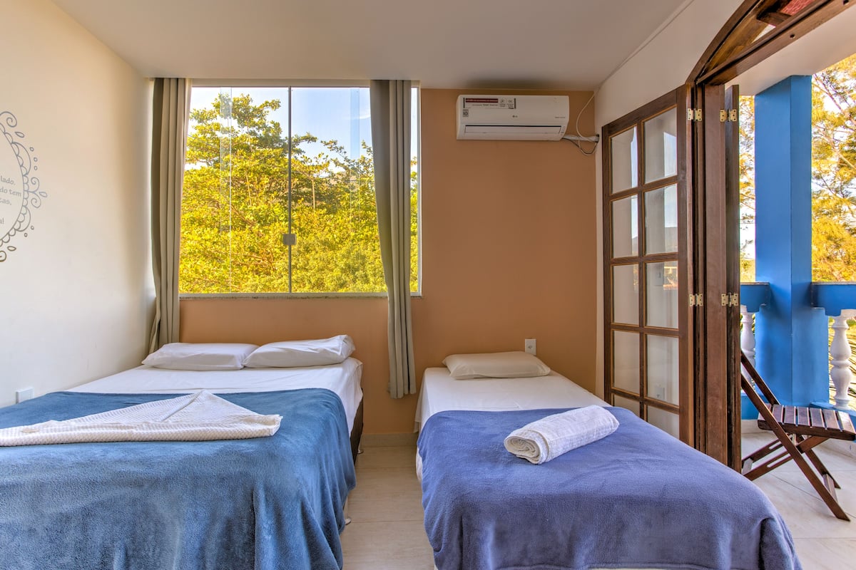 Quarto com duas camas, janela com cortinas, ar condicionado e vista para árvores.