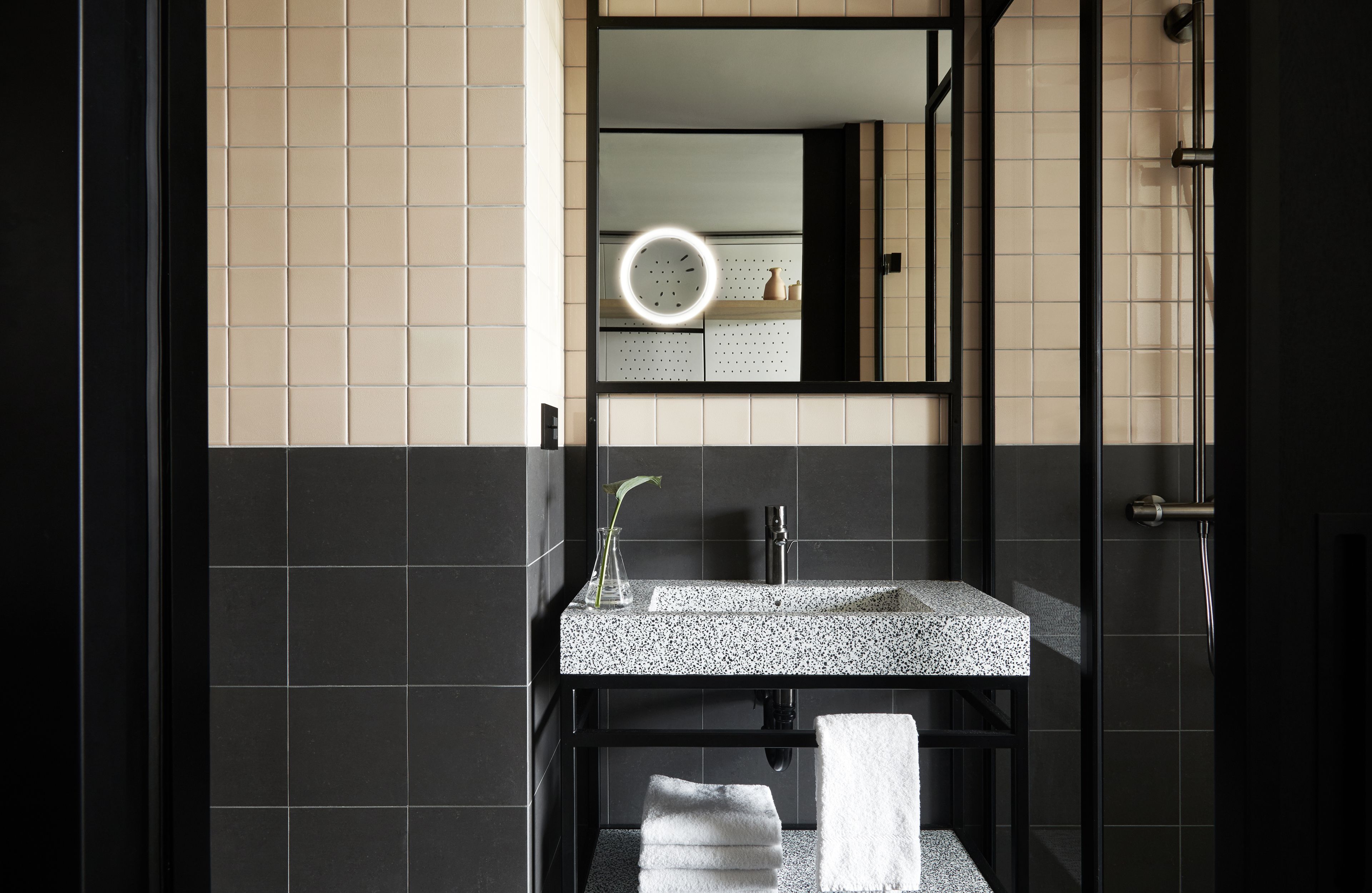 Um banheiro moderno com parede revestida em azulejos preto e bege, pia com bancada de granito, espelho com iluminação circular e porta-toalhas.