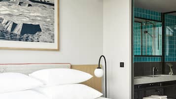 Quarto de hotel moderno com uma cama, almofadas brancas, uma obra de arte emoldurada, um abajur, um banheiro com parede revestida de azulejos azuis, uma pia e um porta-toalhas.