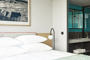 Quarto de hotel moderno com uma cama, almofadas brancas, uma obra de arte emoldurada, um abajur, um banheiro com parede revestida de azulejos azuis, uma pia e um porta-toalhas.