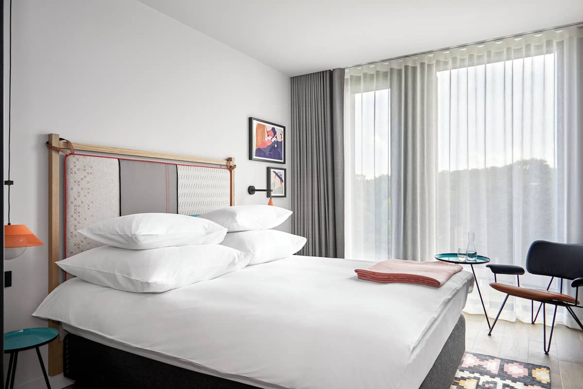 Quarto de hotel com cama, uma cadeira, uma mesinha e uma janela com cortinas.