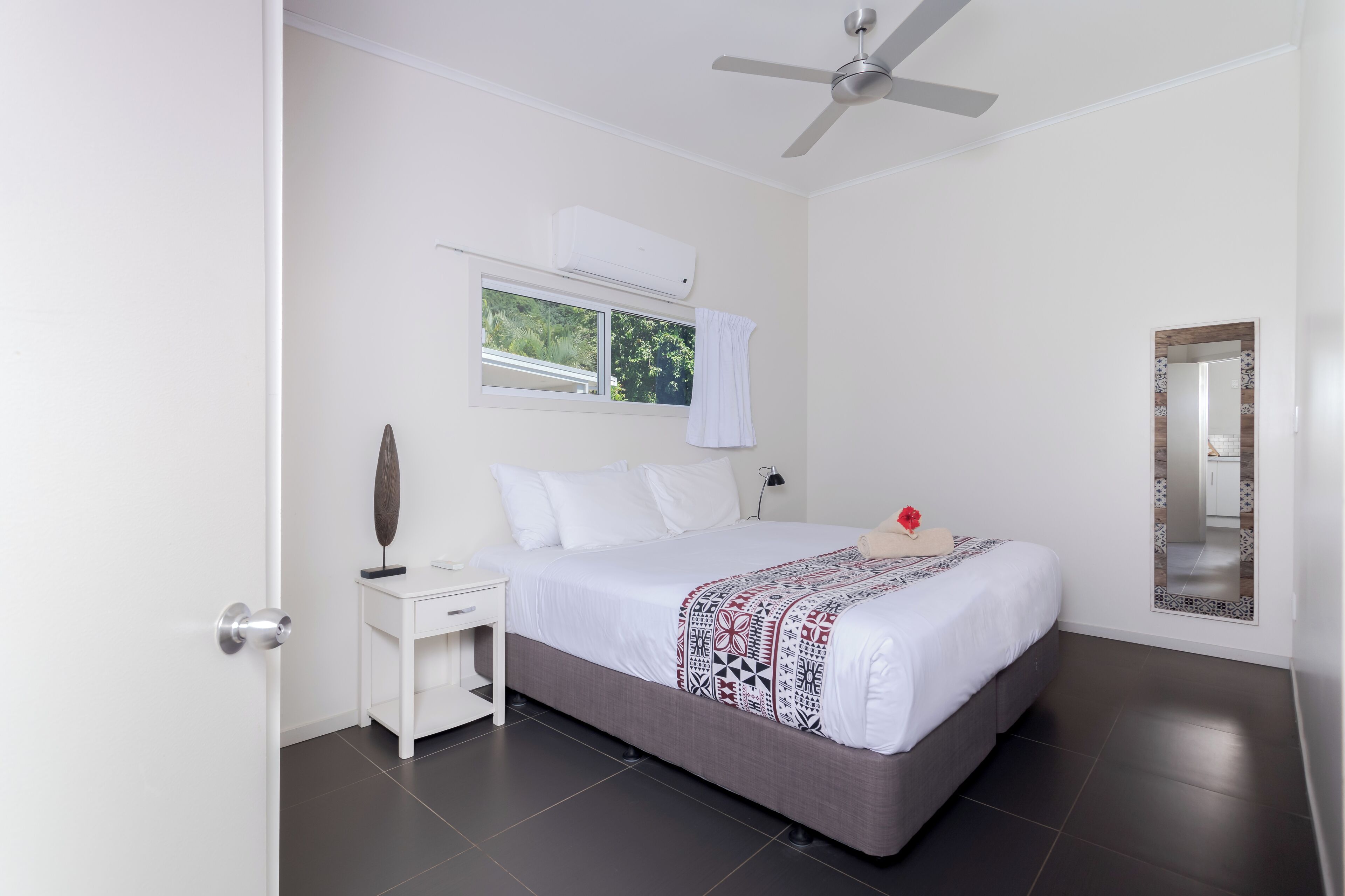 Quarto com cama, mesa de cabeceira, janela com cortinas, ventilador de teto e espelho.