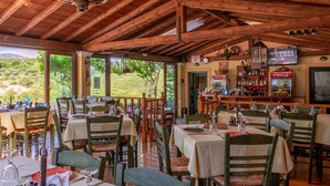 Um restaurante com vigas de madeira, mesas postas com toalhas brancas e um bar com várias garrafas e uma tela de televisão.