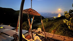 Uma cadeira balanço com assento trançado e estrutura metálica, com vista para uma paisagem costeira onde se vê o mar e luzes distantes da cidade.