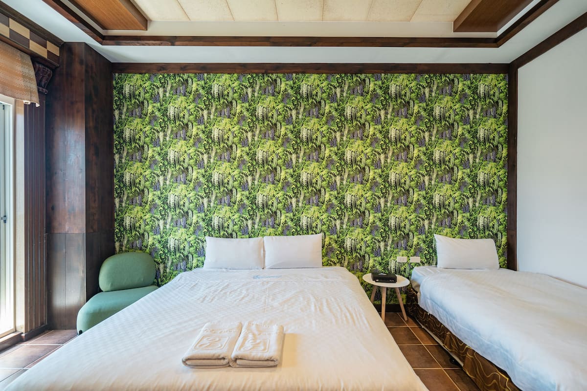 Quarto de hotel com uma cama grande, duas almofadas, uma cadeira verde, uma mesa redonda pequena e papel de parede estampado.