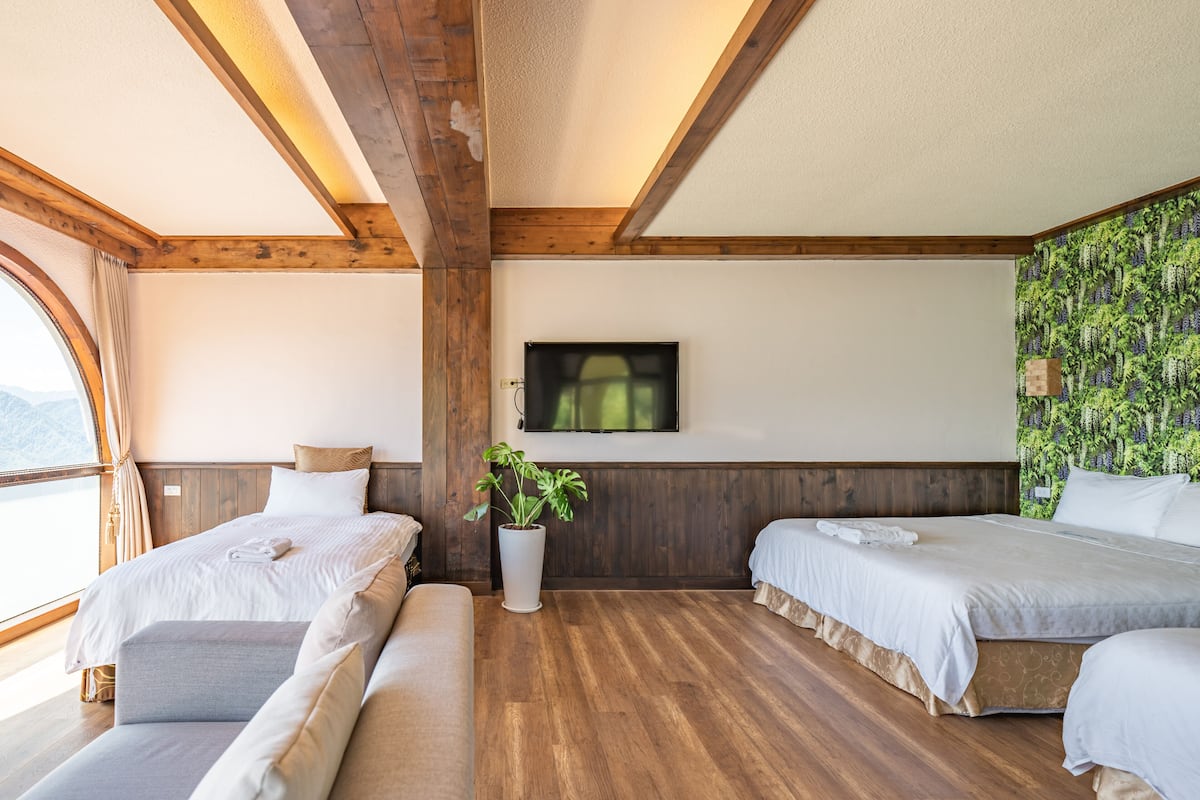 Quarto com duas camas, uma janela ampla, vigas de madeira, uma planta em vaso e uma televisão de tela plana na parede.