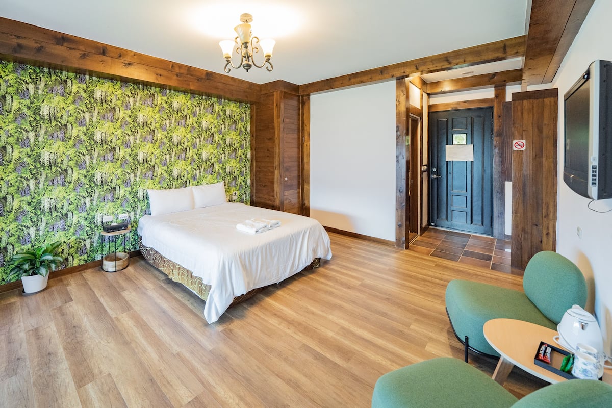 Um quarto com uma cama, uma poltrona verde, uma mesinha com chaleira e uma televisão fixada na parede.