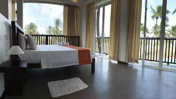 Quarto de hotel com cama, mesa de cabeceira, abajur, tapete branco e janelas grandes com cortinas.