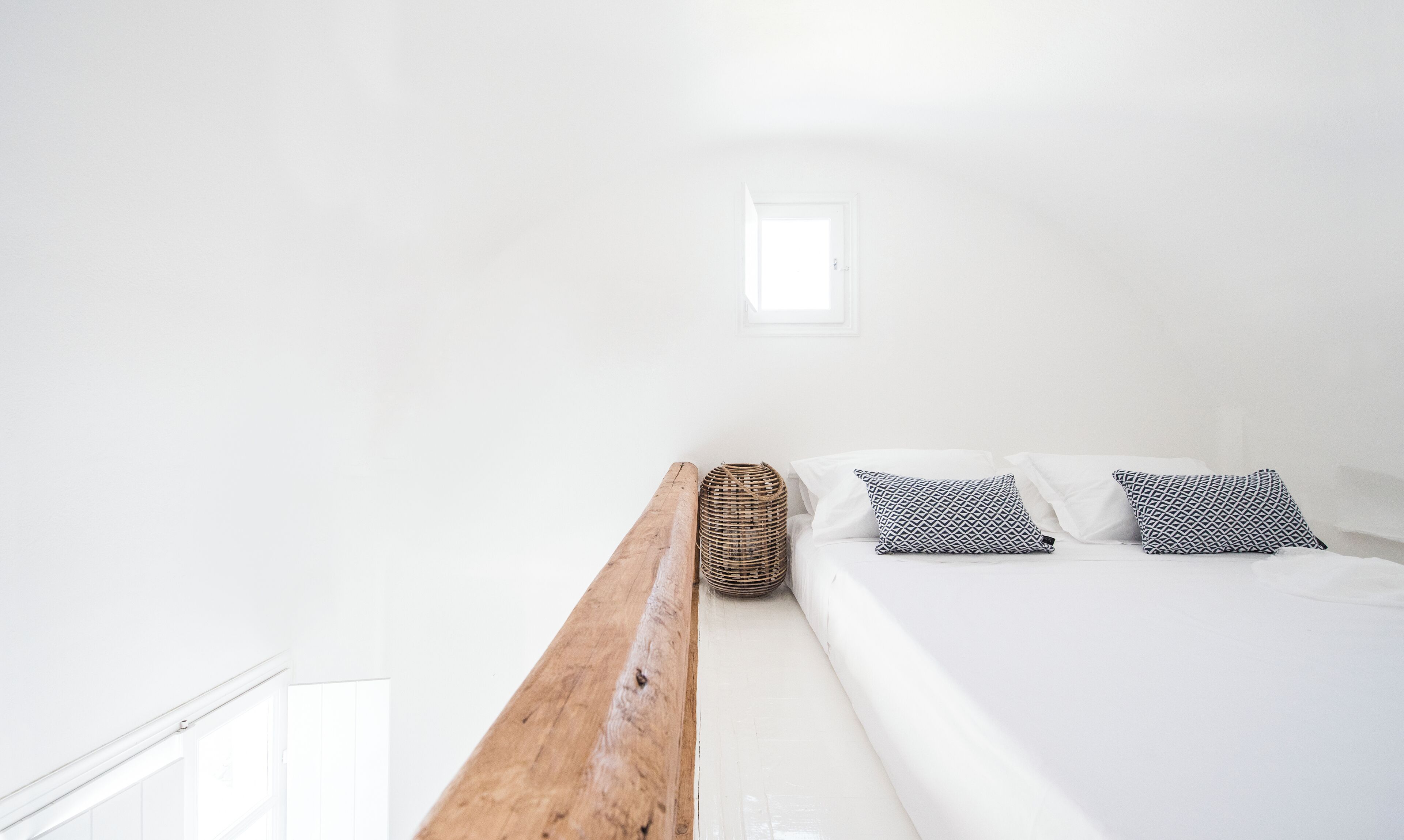 Quarto minimalista com cabeceira de madeira, uma pequena janela e cama com roupa de cama branca e almofadas estampadas.