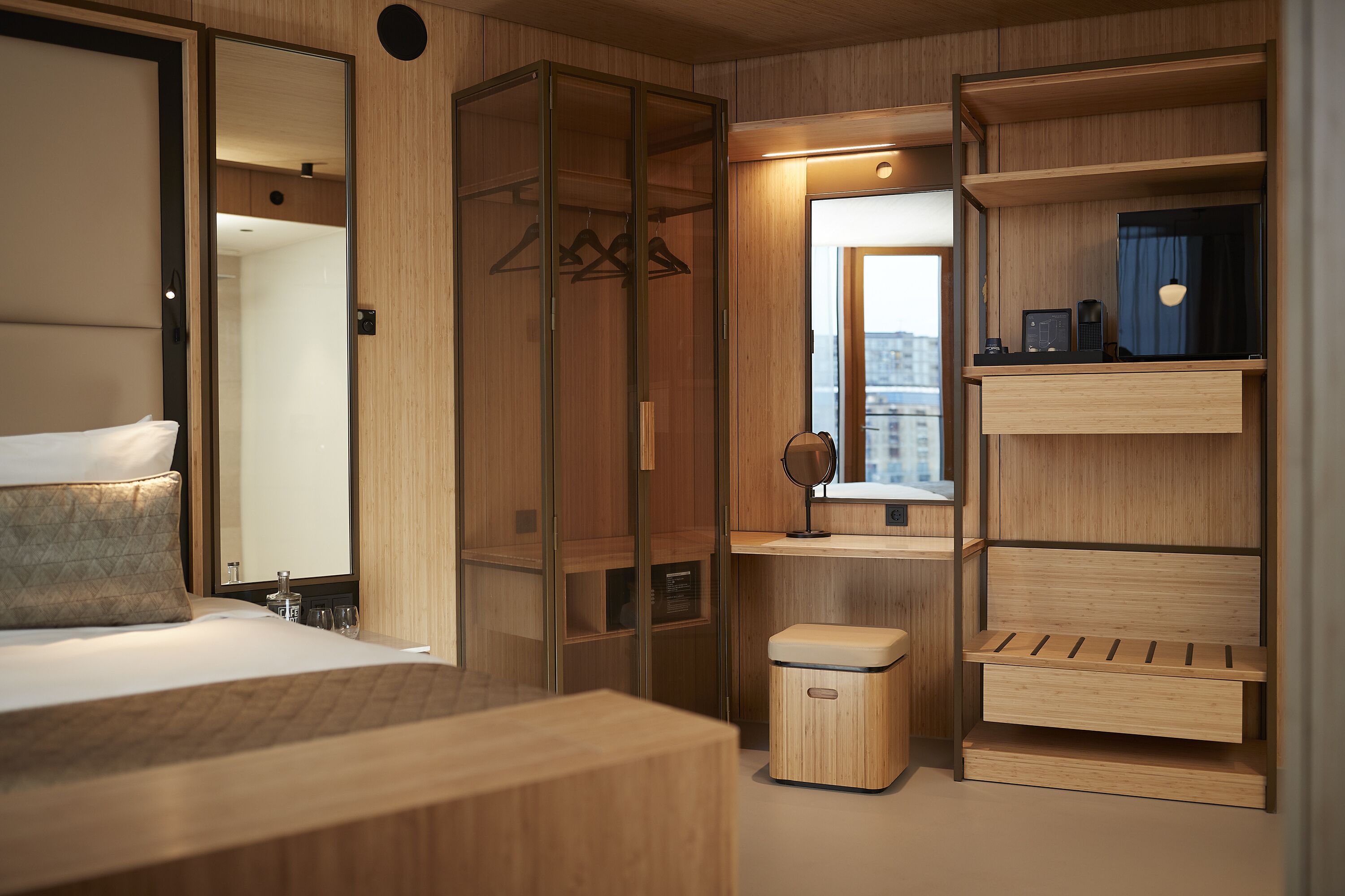 Quarto de hotel moderno com cama, guarda-roupa de madeira, um banquinho e um espelho.