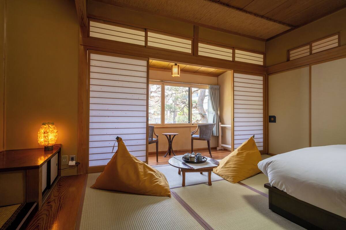 Um quarto tradicional em estilo japonês, com tatames, uma cama, uma pequena mesa com um tabuleiro e uma cadeira.