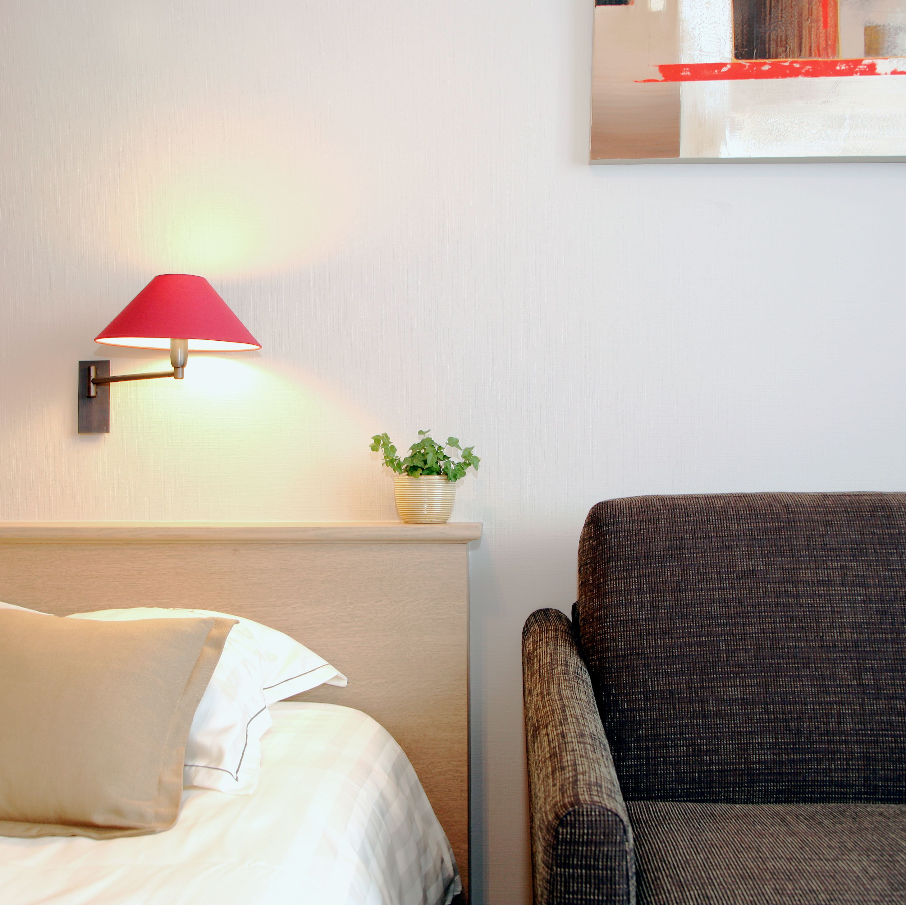 Quarto de hotel com uma cama, um abajur vermelho, uma pequena planta em vaso e uma poltrona escura.