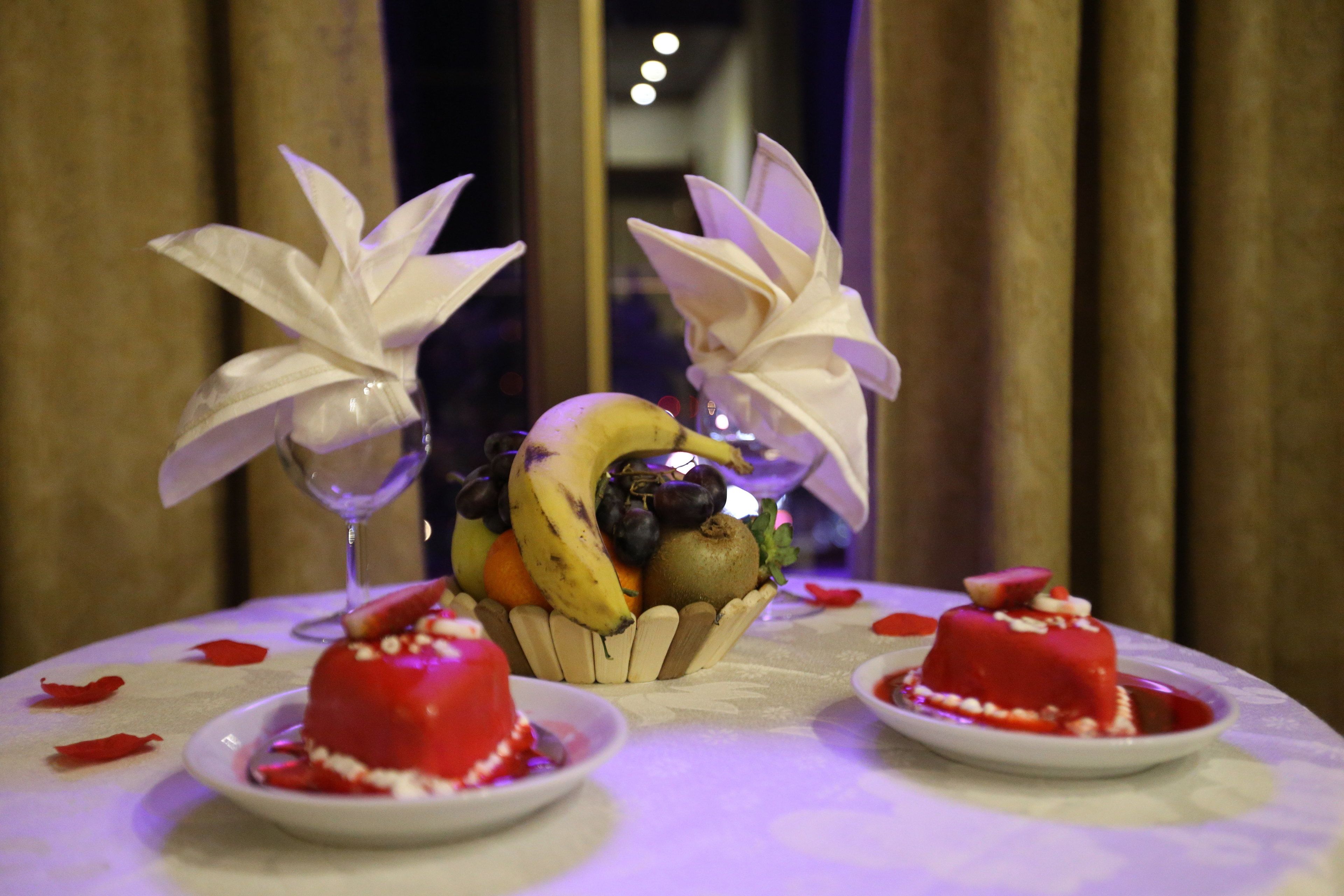 Uma mesa com uma composição de frutas, duas decorações florais brancas e dois doces vermelhos.