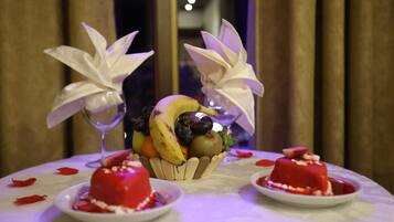 Uma mesa com uma composição de frutas, duas decorações florais brancas e dois doces vermelhos.