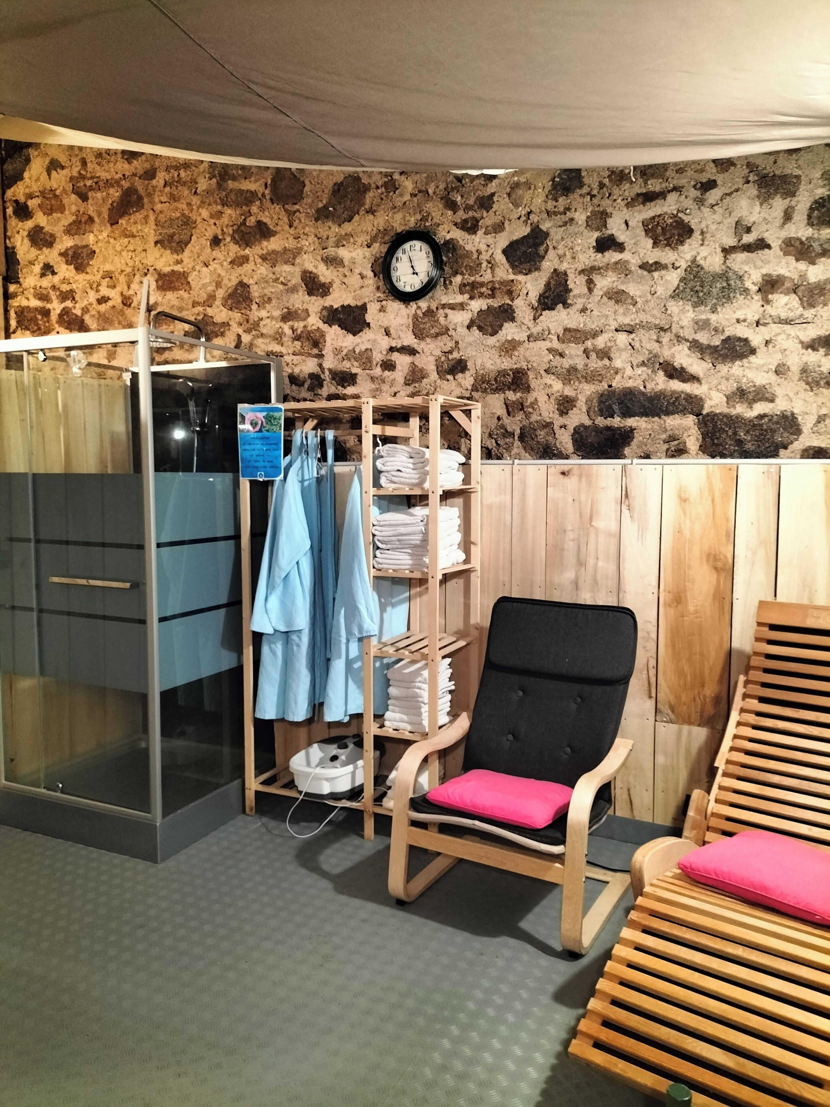 Quarto com parede de pedra, um relógio, uma cadeira com almofada rosa, uma prateleira de madeira com toalhas e uma porta de vidro.