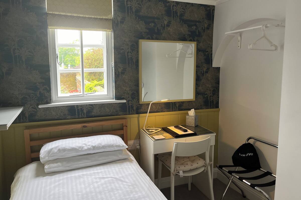 Quarto com cama, escrivaninha, cadeira, espelho, janela com vista para vegetação e um cabideiro.