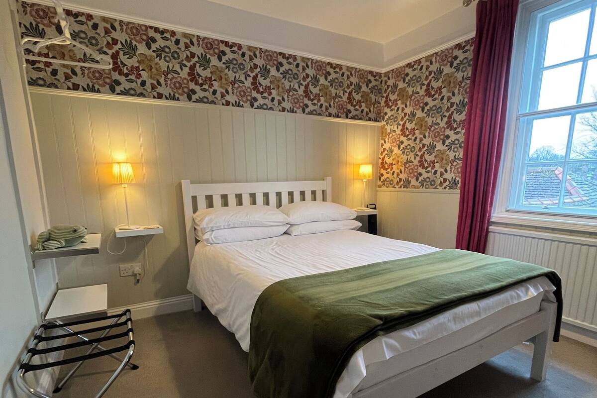 Quarto bem arrumado com cama, luminárias de cabeceira, radiador e janela com cortinas.