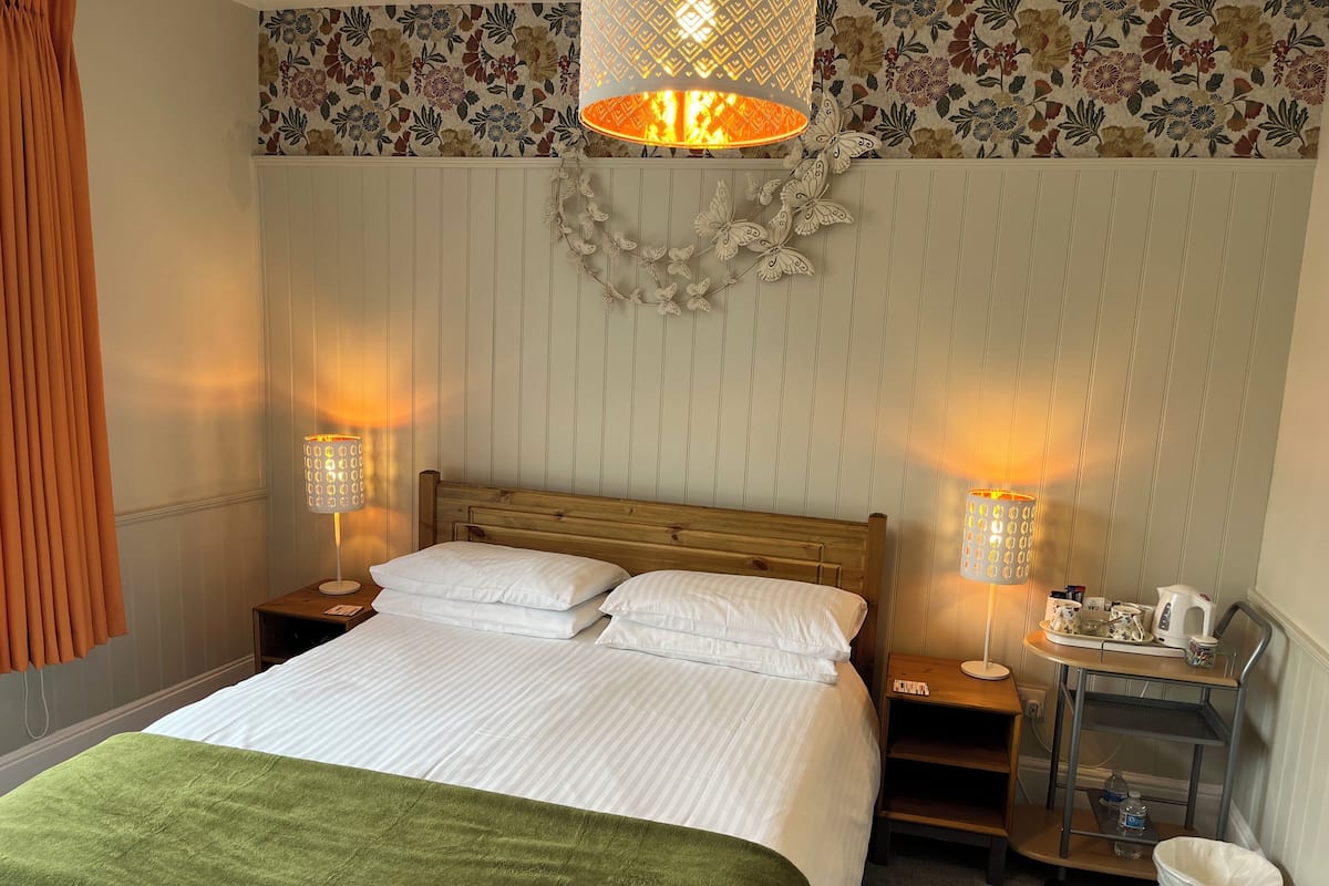 Cama bem arrumada com lençóis brancos, duas luminárias de cabeceira, um painel de cabeceira de madeira, um cobertor verde e uma parede com padrão floral, além de uma pequena mesa com chaleira e xícaras.