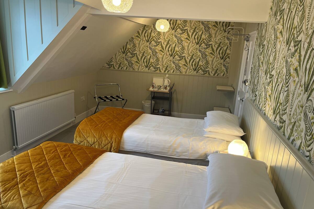 Quarto de hotel com duas camas, um radiador, uma mesinha com uma chaleira e uma cadeira.