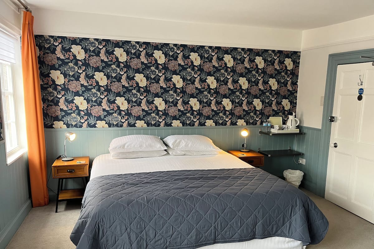 Quarto com cama, duas mesas de cabeceira, um criado-mudo, papel de parede floral e uma porta.