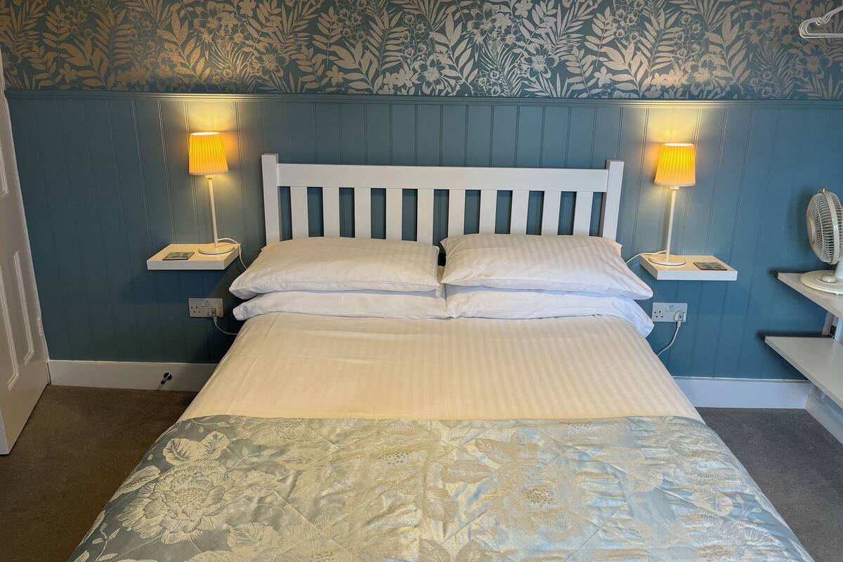 Cama bem arrumada com roupa de cama branca, duas luminárias de cabeceira e um ventilador fixado na parede.