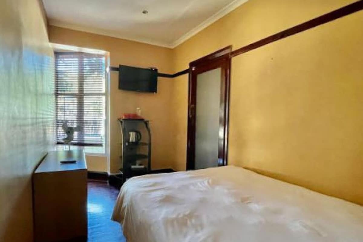 Quarto de hotel com cama, escrivaninha, televisão fixada na parede e janela com persianas.
