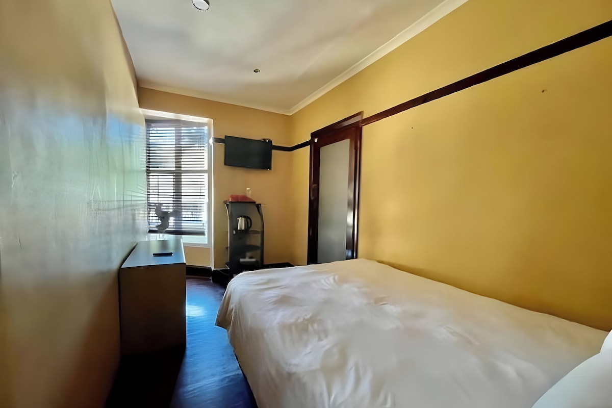 Quarto de hotel com cama, escrivaninha, cadeira, TV fixada na parede e janela com persianas.
