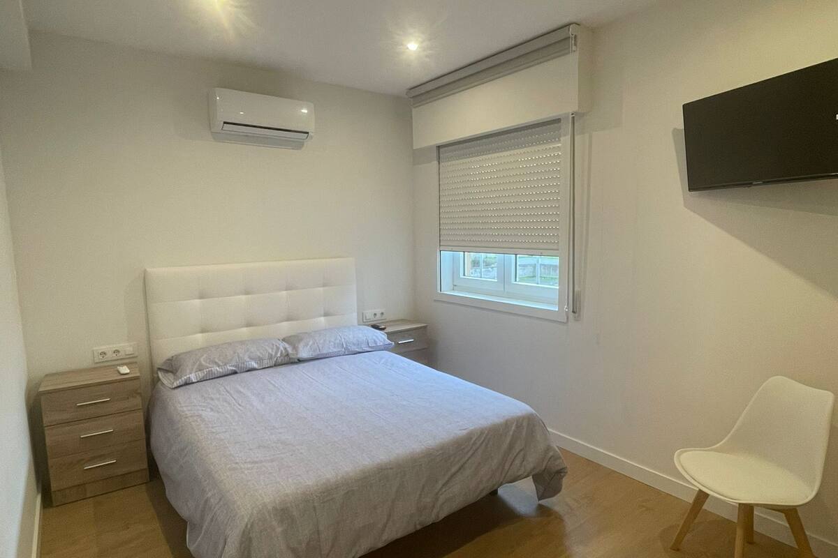 Quarto com cama, mesa de cabeceira, cadeira, ar condicionado, janela com persianas e televisão na parede.