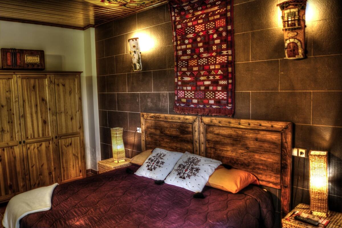 Quarto com cabeceira de madeira, cama grande com colcha vermelha, duas luminárias fixadas na parede e tapeçaria acolchoada acima da cama.