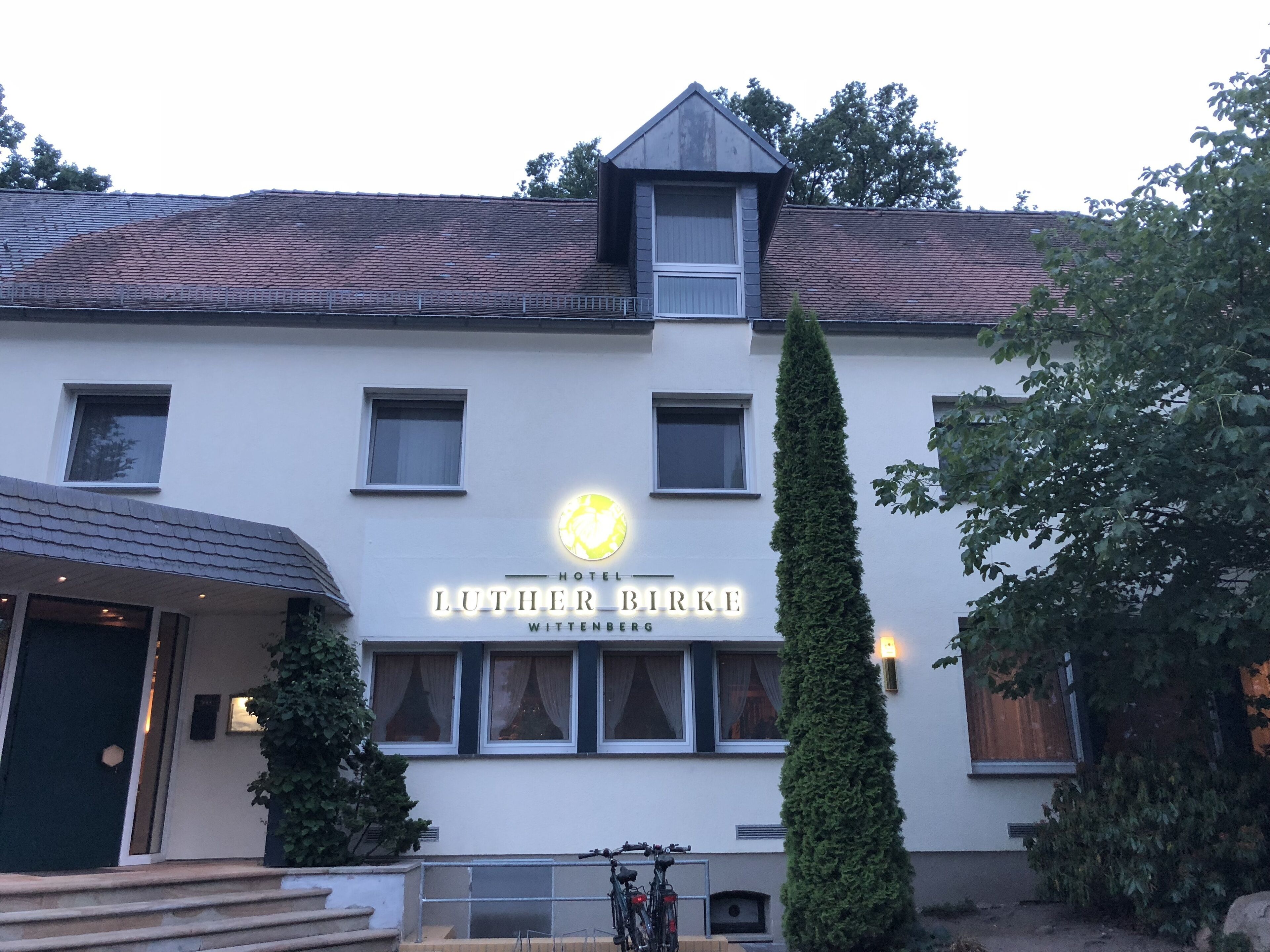 Um hotel chamado 'LUTHER BIRKE' com arquitetura europeia tradicional, apresentando uma entrada imponente, uma árvore conífera alta e uma bicicleta estacionada na frente.