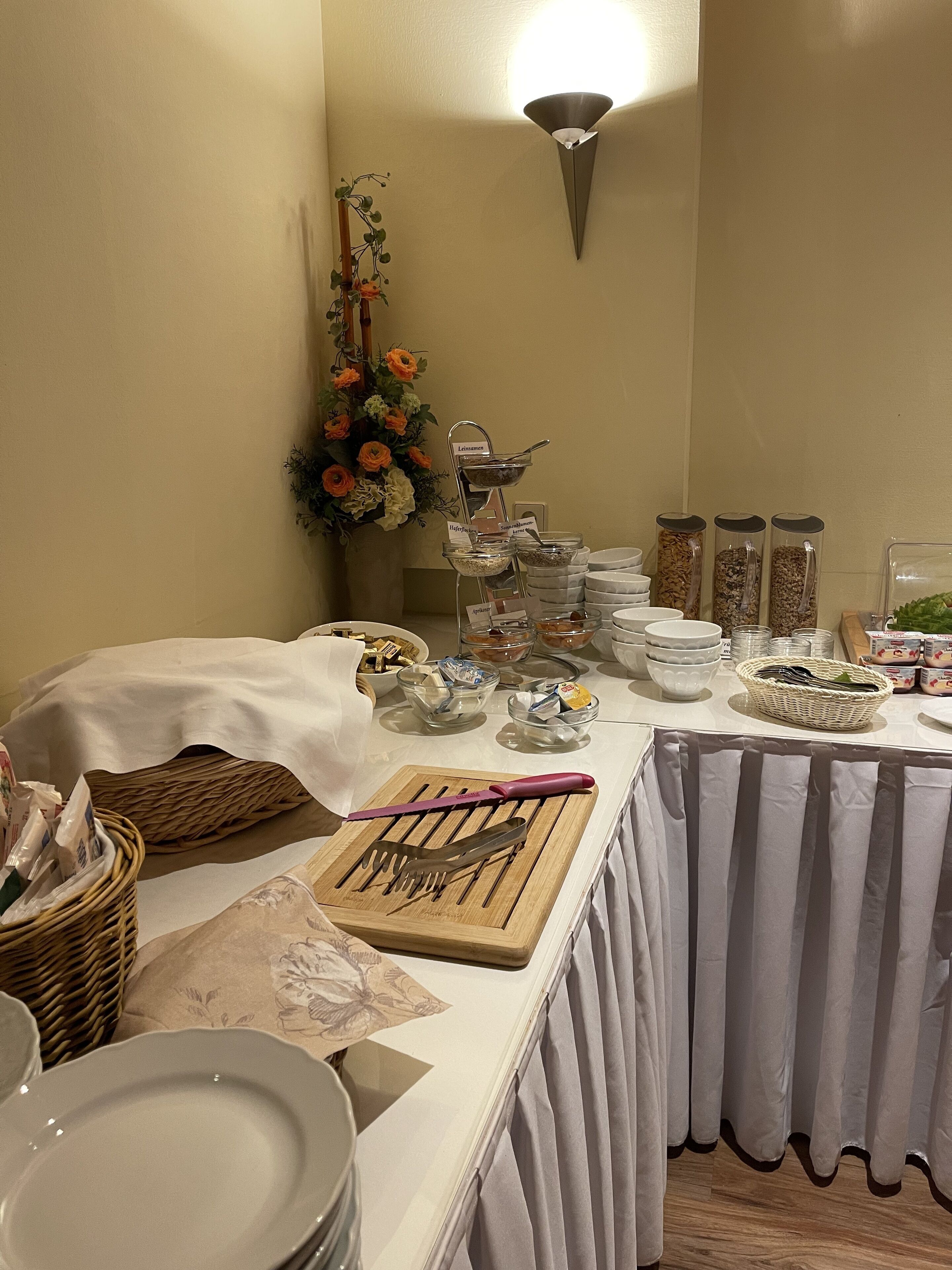 Disposição de buffet com diversos recipientes de comida, uma faca e colher sobre uma tábua de corte, e um arranjo floral na parede.