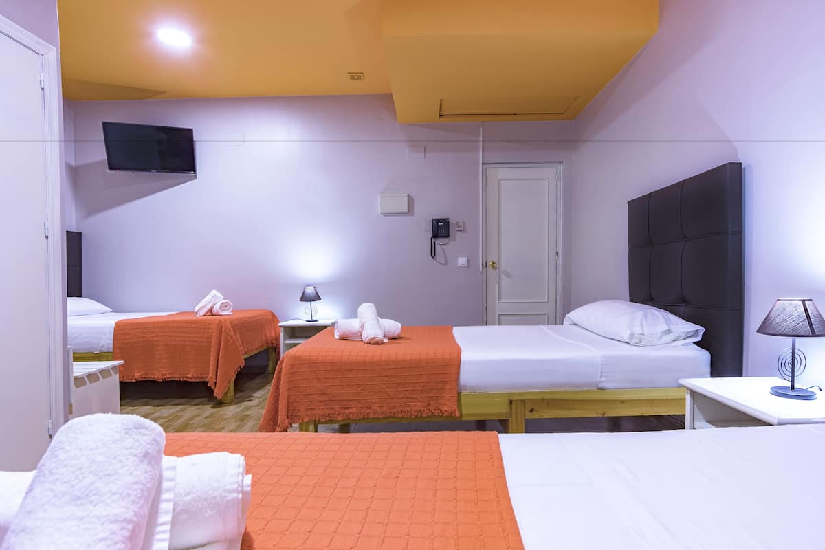 Quarto de hotel com duas camas de solteiro, uma televisão na parede, uma mesa de cabeceira com abajur e uma porta.