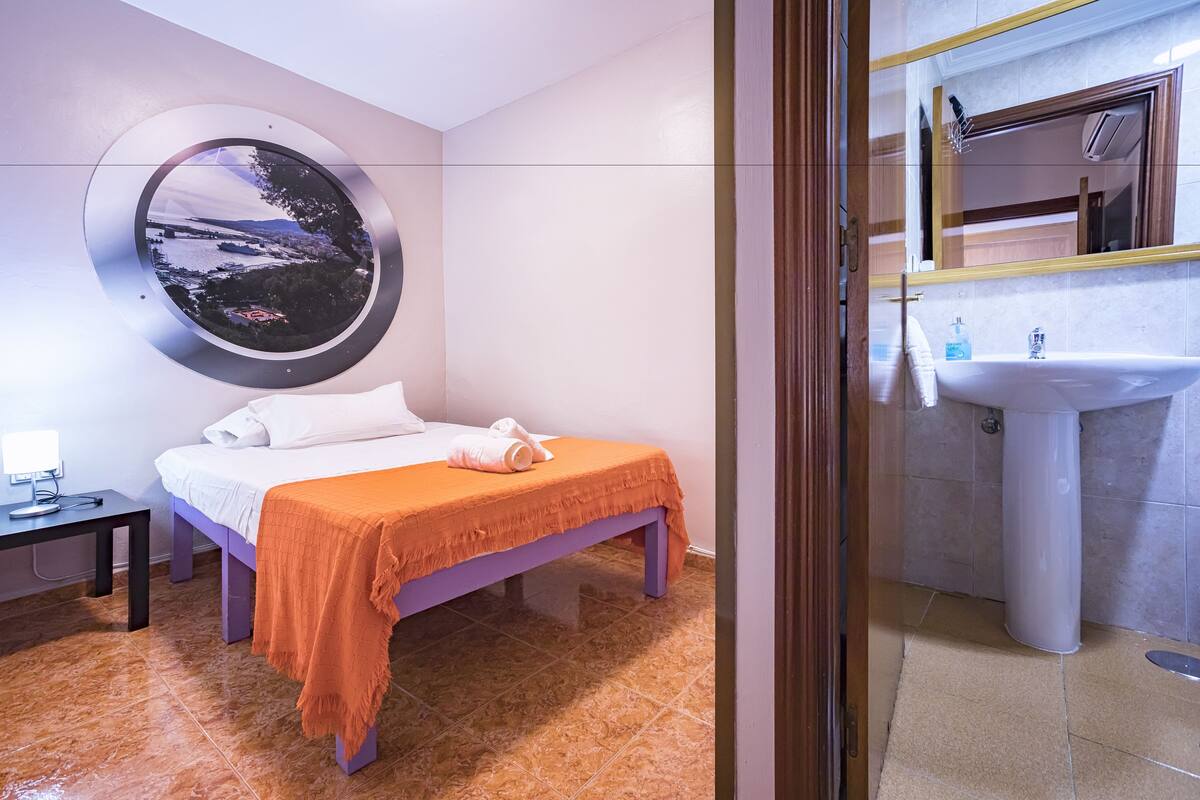 Quarto com cama, mesa de cabeceira, espelho redondo e vista para uma paisagem pela janela circular.