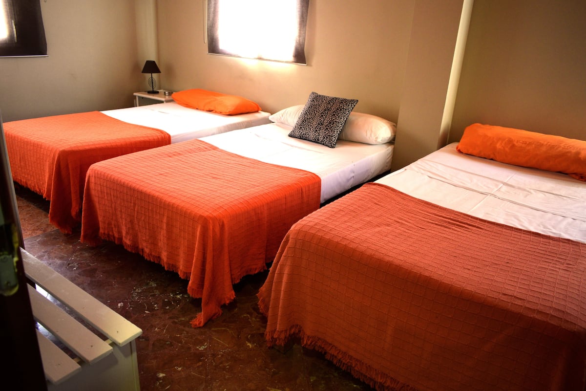 Quarto de hotel com três camas de solteiro, cada uma com colcha laranja e roupa de cama branca. Há uma pequena mesa com uma luminária e uma janela acima das camas.