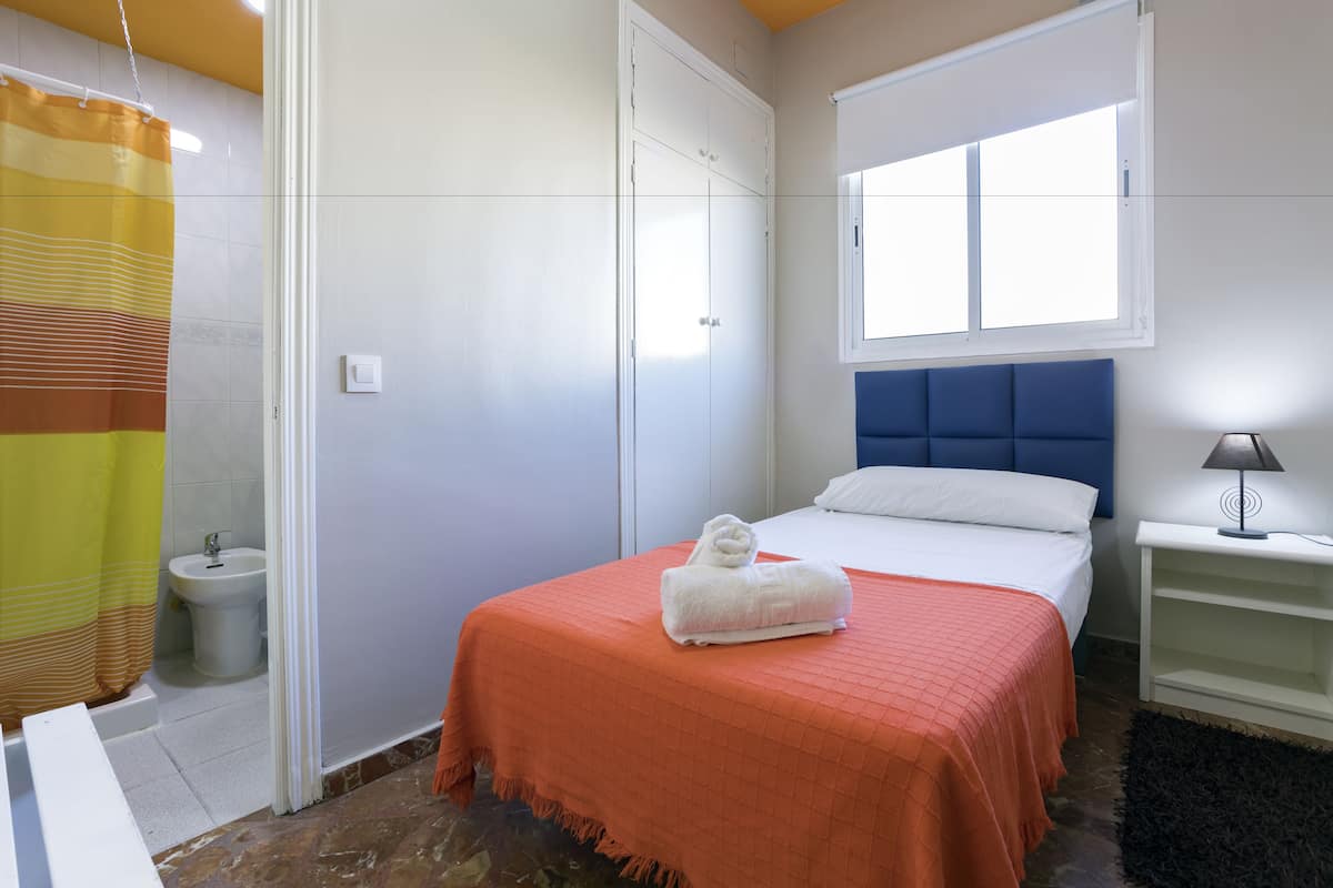 Quarto com cama, mesa de cabeceira, janela e banheiro com cortina de chuveiro.