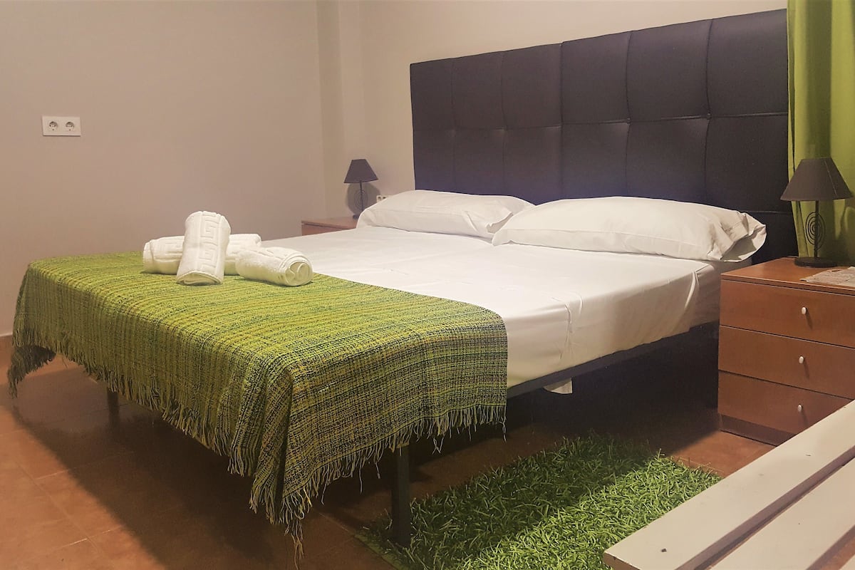 Uma cama bem arrumada com lençóis brancos, um cobertor verde com textura e toalhas brancas dobradas em uma mesinha de cabeceira de madeira.