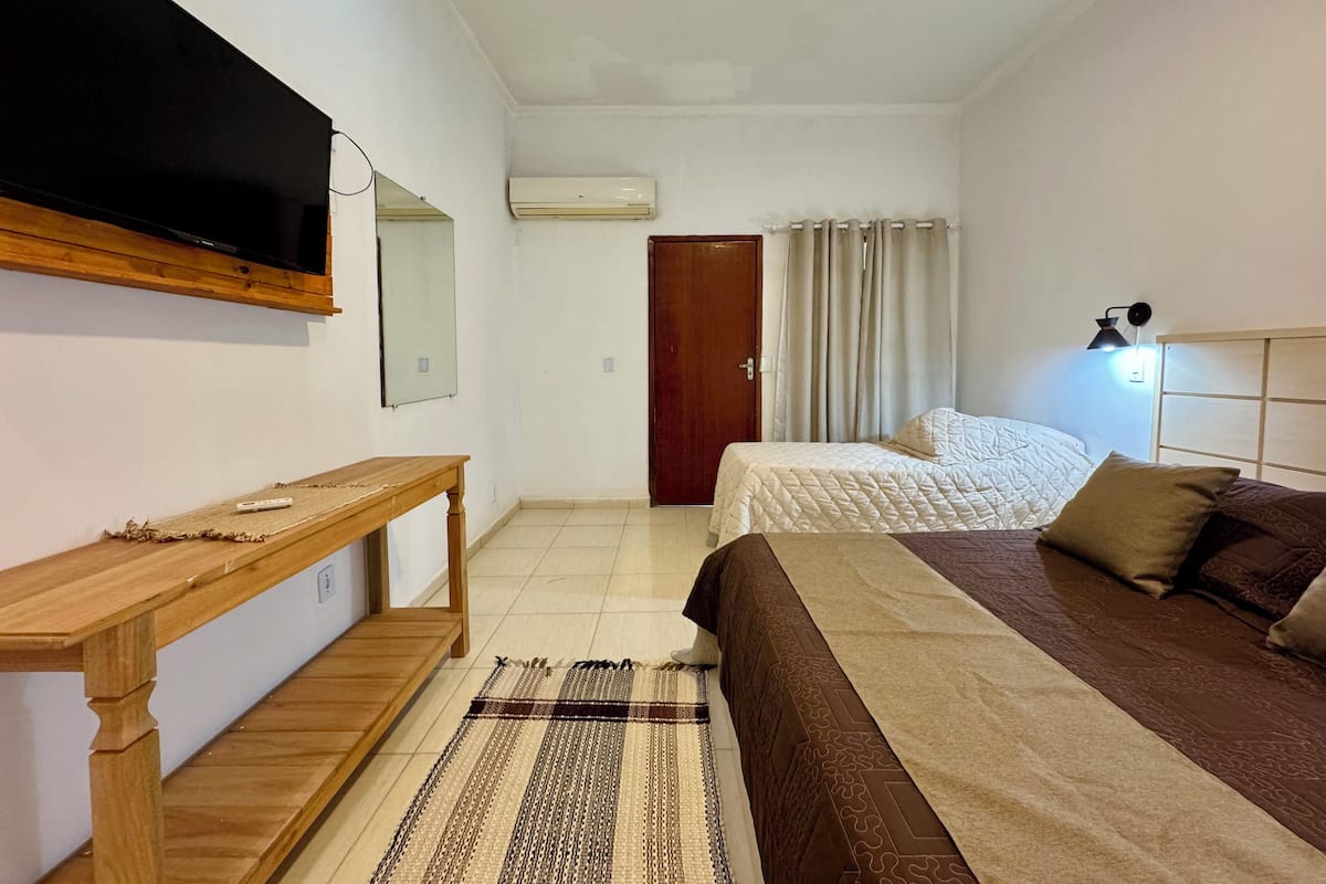 Quarto com cama, televisão, espelho, mesa de madeira, tapete e ar condicionado.