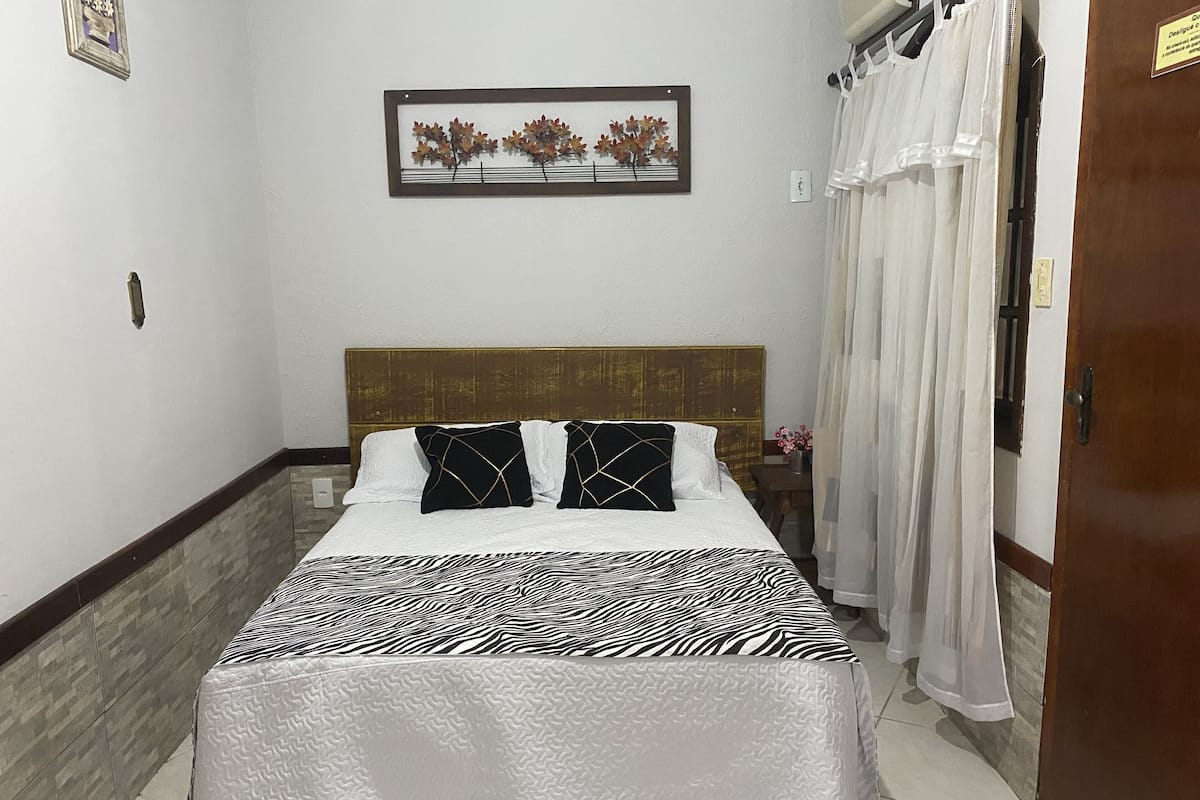 Uma cama bem arrumada com colcha estampada em zebra e duas almofadas decorativas, uma cabeceira de madeira, um quadro na parede e uma porta com cortina.