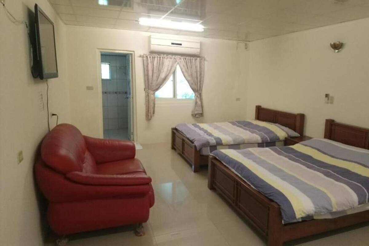 Quarto com duas camas, uma poltrona vermelha, televisão e janela com cortinas.